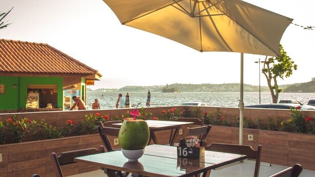 Um restaurante à beira-mar com mesas ao ar livre, um grande guarda-sol e vista para a água.
