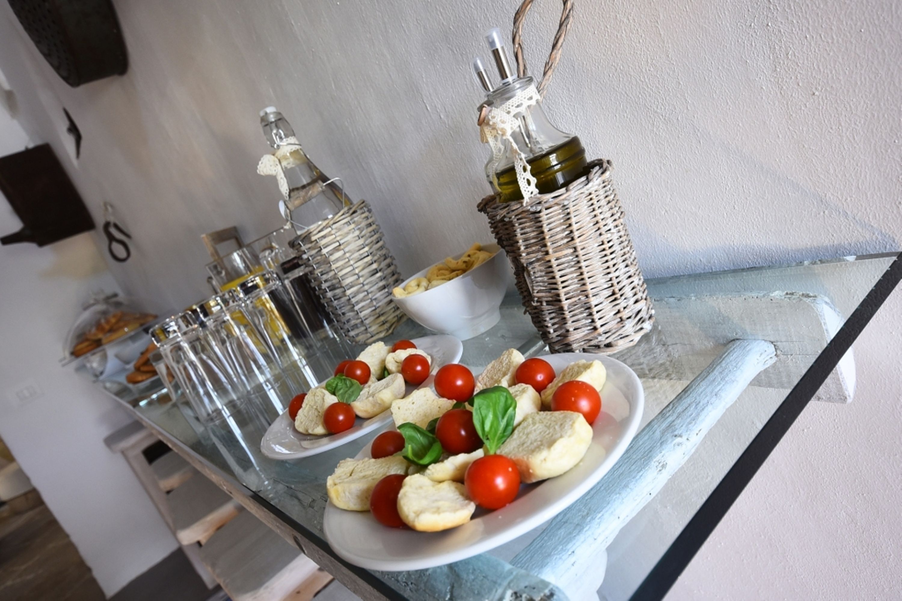Mesa de vidro com pratos de bruschetta e tomates cereja, rodeada por cestos de vime com garrafas de vidro.