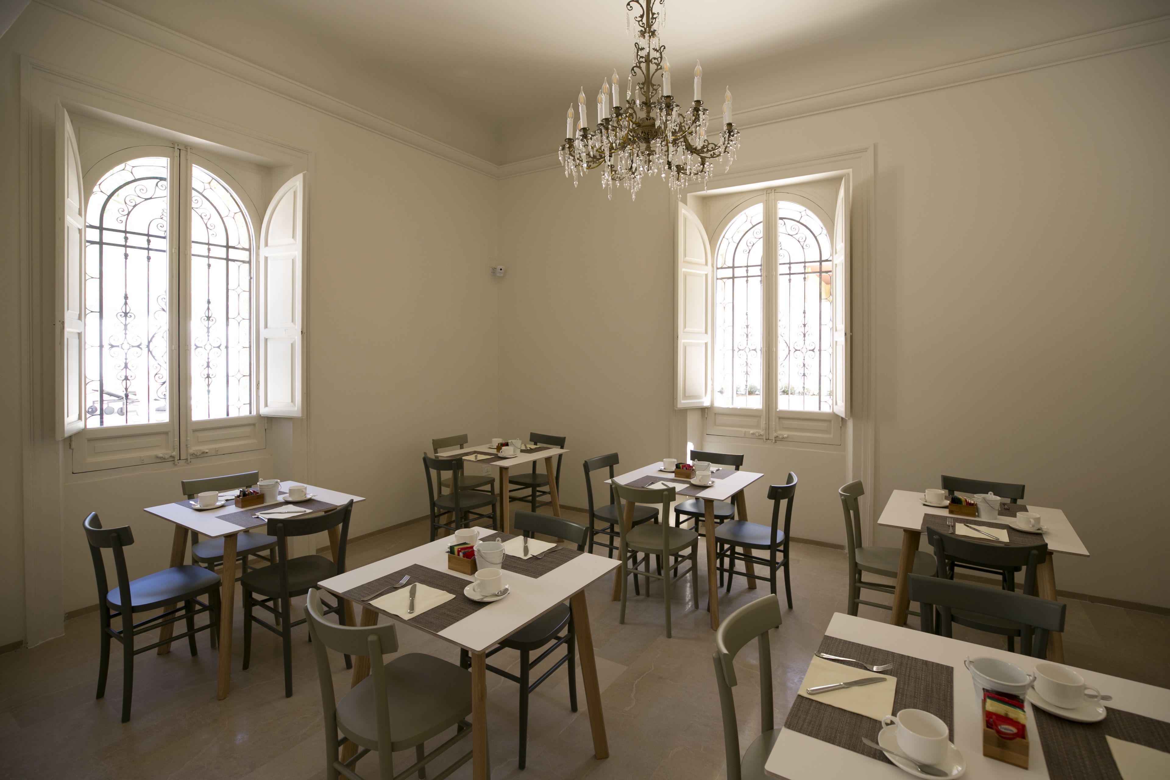 Uma sala de jantar com várias mesas e cadeiras, janelas amplas com detalhe em ferro e um candelabro.