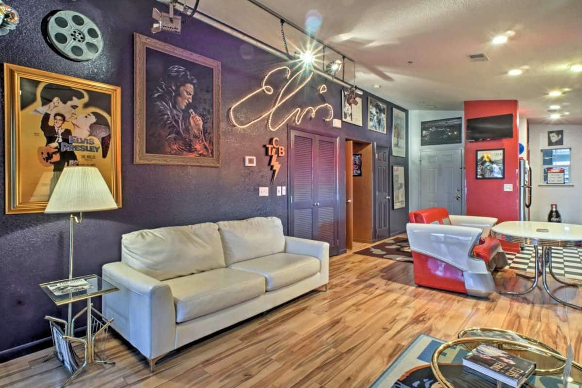 Quarto com sofá branco, mesa de centro de vidro, cadeira vermelha e branca, e um letreiro neon na parede.