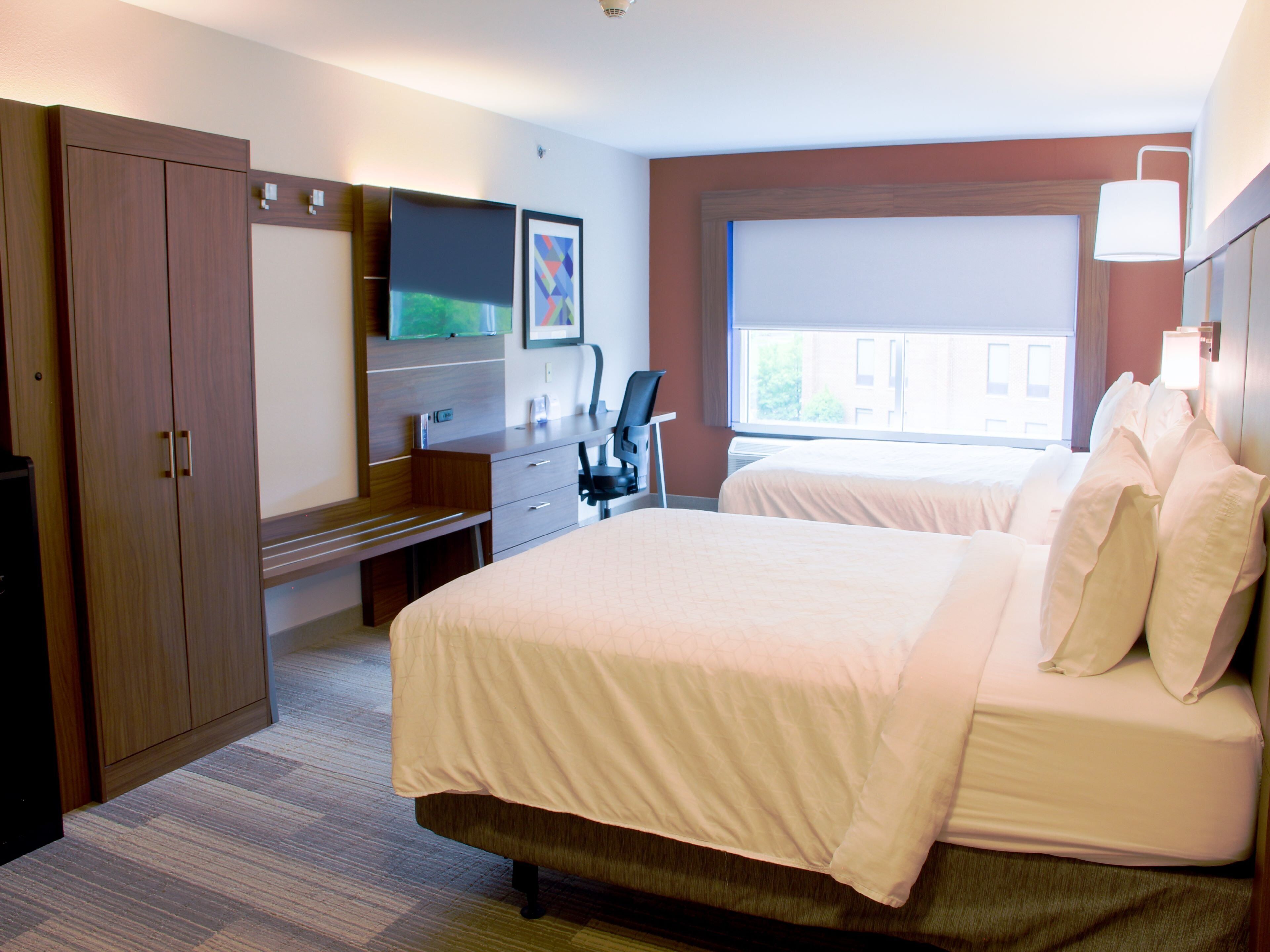 Quarto de hotel com uma cama grande, uma escrivaninha, um guarda-roupa, uma televisão e uma janela com vista para edifícios.