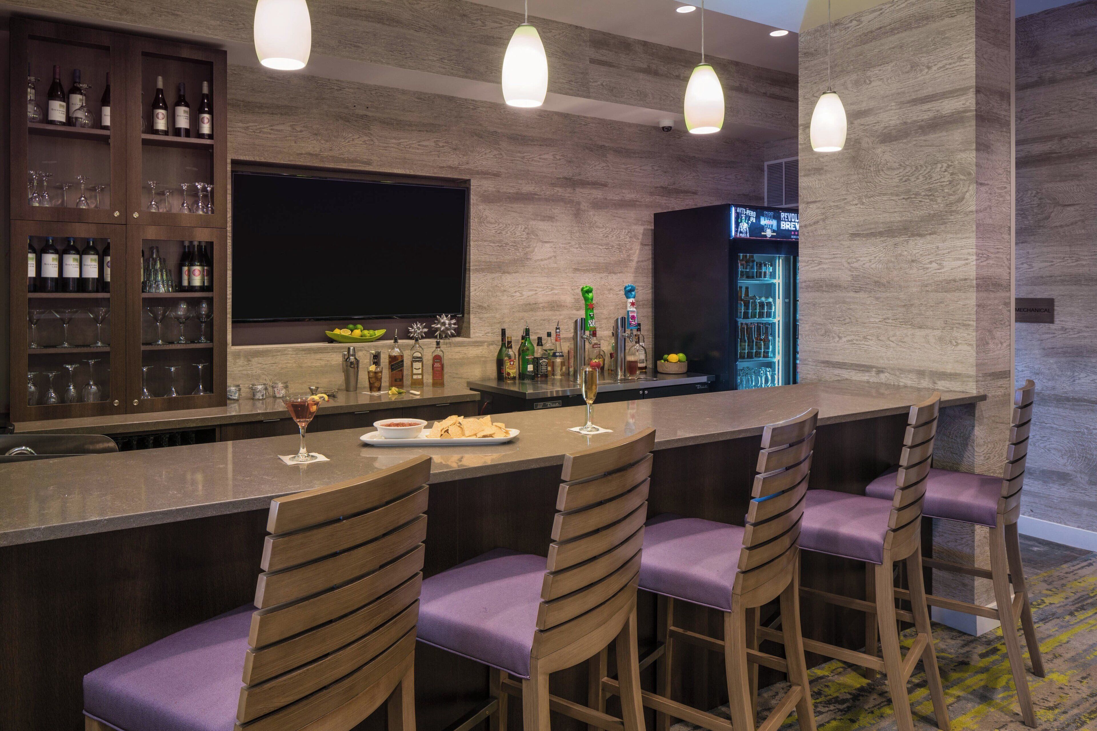 Uma área de bar com um balcão comprido, diversas opções de assento e variedade de garrafas e copos.