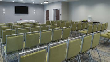Uma sala de conferências com fileiras de cadeiras, uma tela grande, uma mesa com um vaso e um bebedouro.