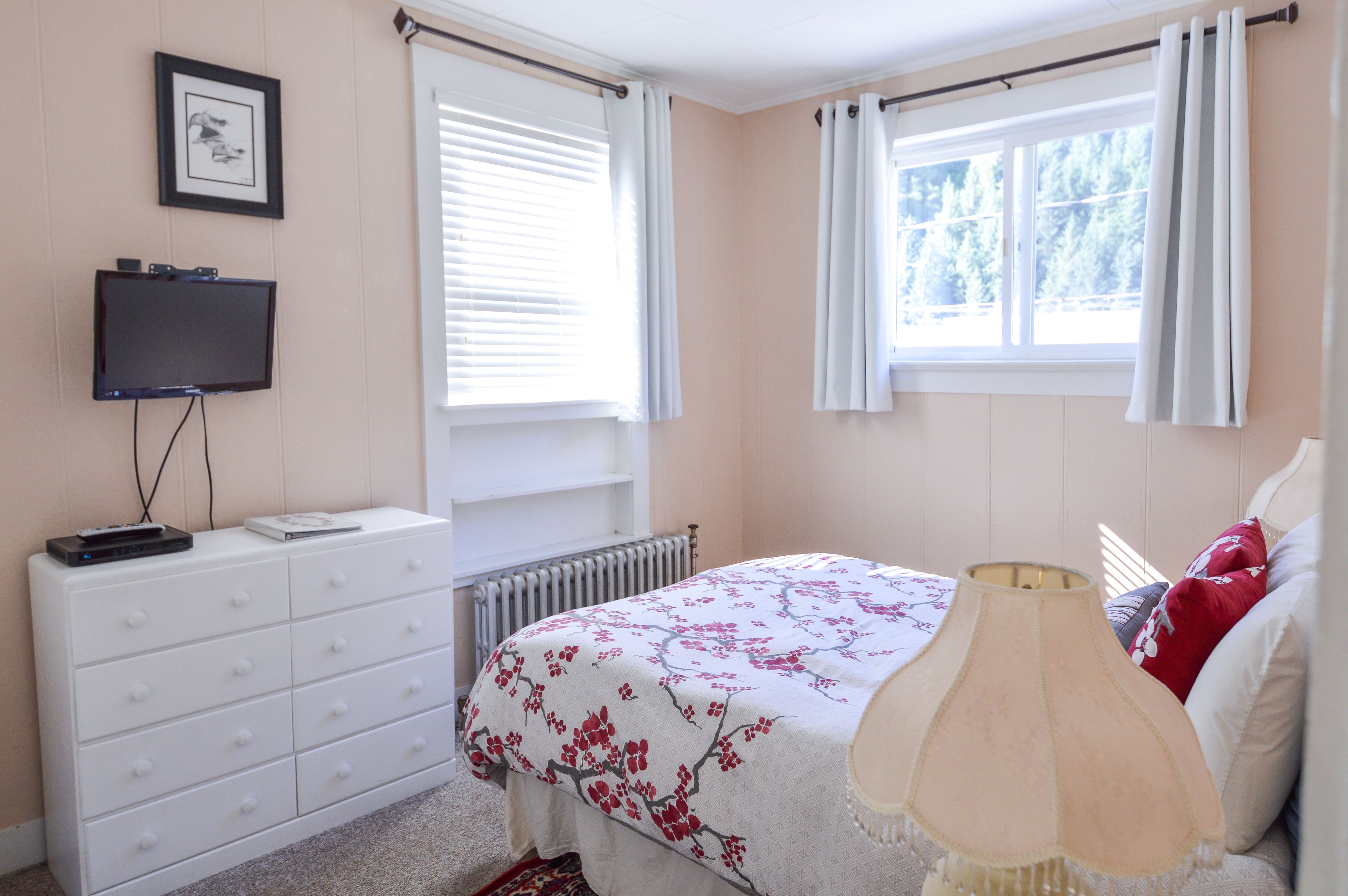 Quarto com cama, cômoda, televisão e janelas com cortinas.