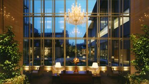 Um saguão moderno com grandes janelas, um candelabro e uma área de estar com mesas e luminárias.
