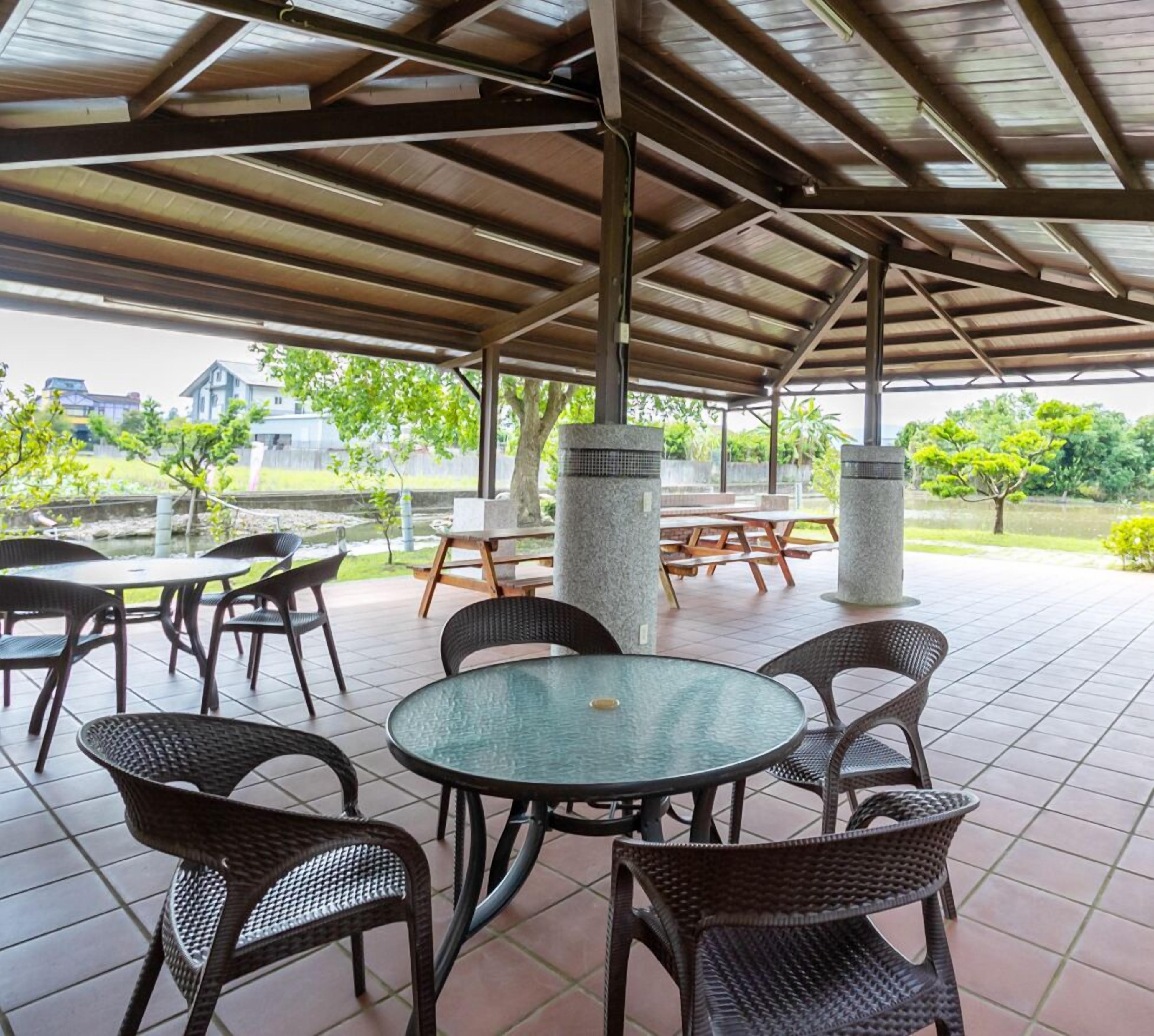 Uma área de estar ao ar livre com mesas redondas e cadeiras de vime, com teto coberto e vista para vegetação e edifícios ao fundo.