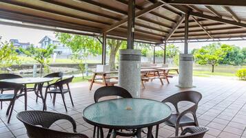 Uma área de estar ao ar livre com mesas redondas e cadeiras de vime, com teto coberto e vista para vegetação e edifícios ao fundo.
