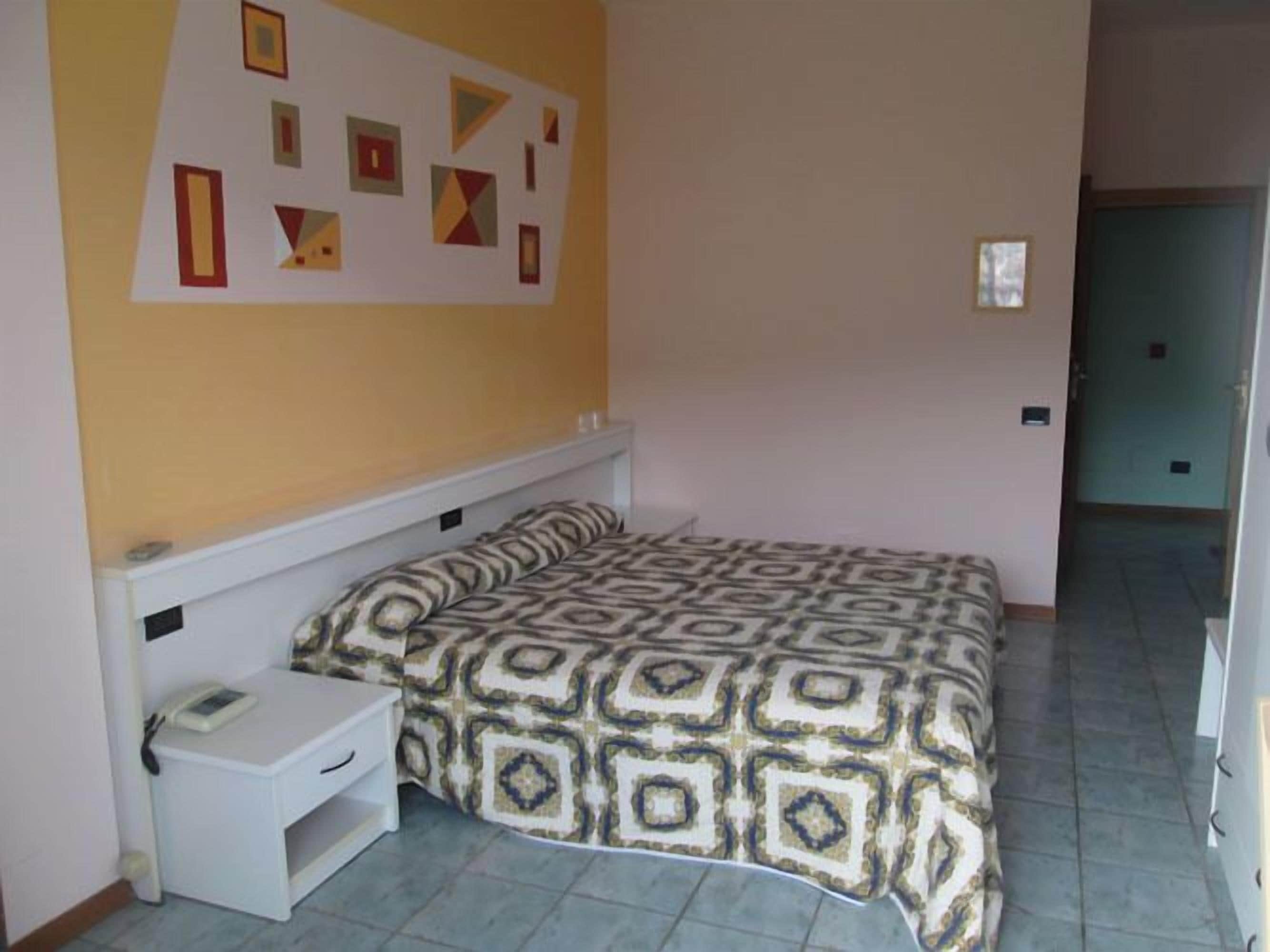 Quarto com cama, mesa de cabeceira com telefone e parede decorada com arte geométrica.