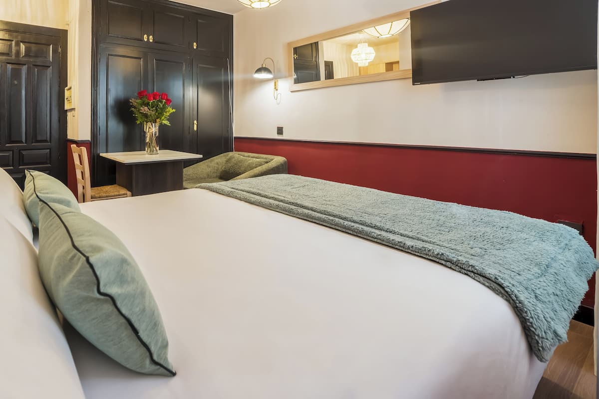 Quarto de hotel com uma cama grande, uma pequena mesa com um vaso de flores, uma cadeira e uma televisão na parede.