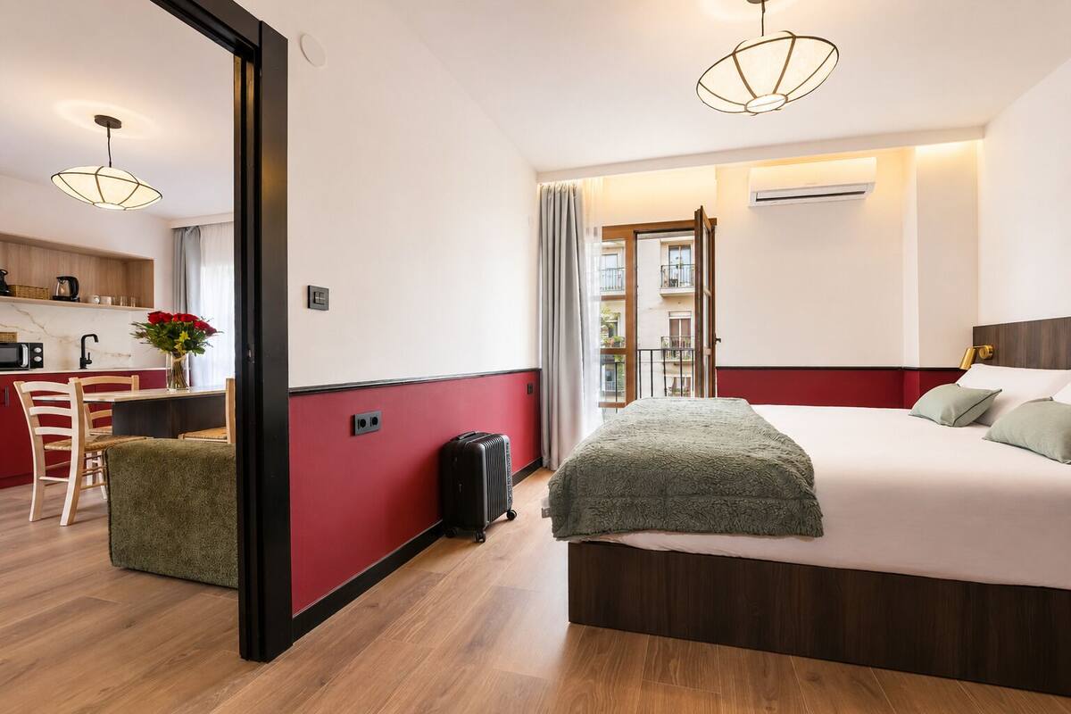 Quarto de hotel com cama, mala, cozinha compacta e vista para o exterior.