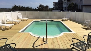 Um deck de madeira com piscina retangular, cadeiras de descanso e uma cerca branca.