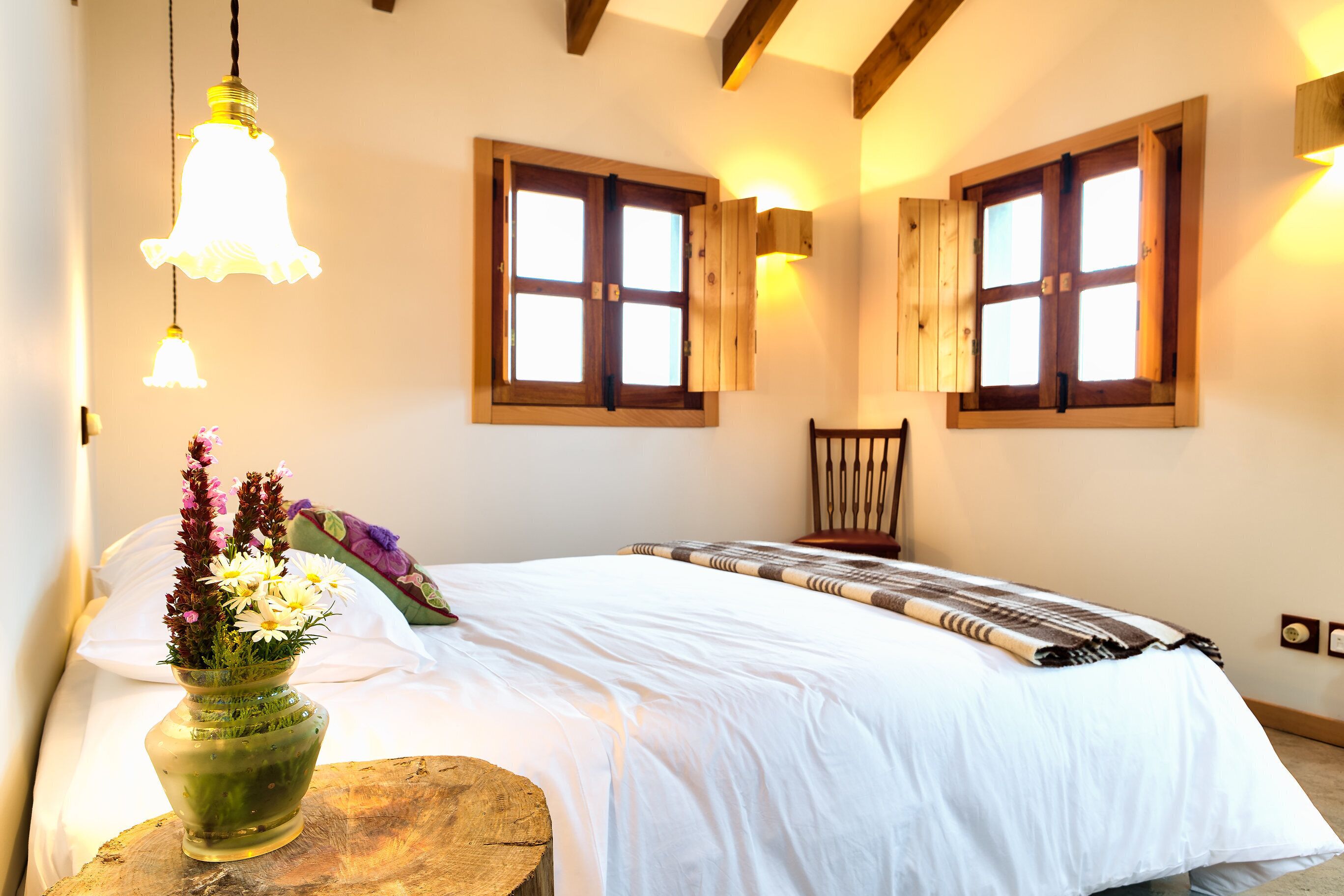 Quarto com cama, uma cadeira de madeira, um vaso com flores e duas janelas com molduras de madeira.
