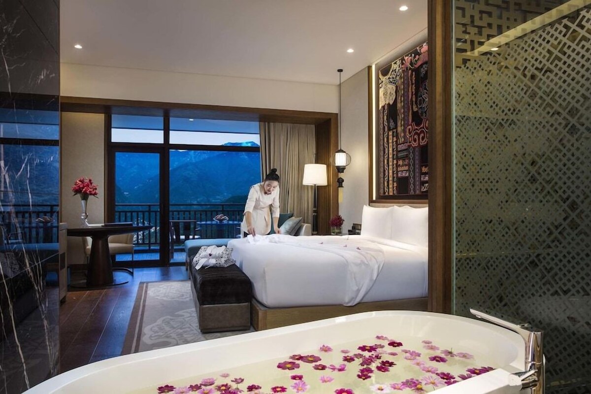 Quarto de hotel com uma cama grande, área de estar com mesa e cadeiras, varanda com vista e banheira com pétalas de flores.