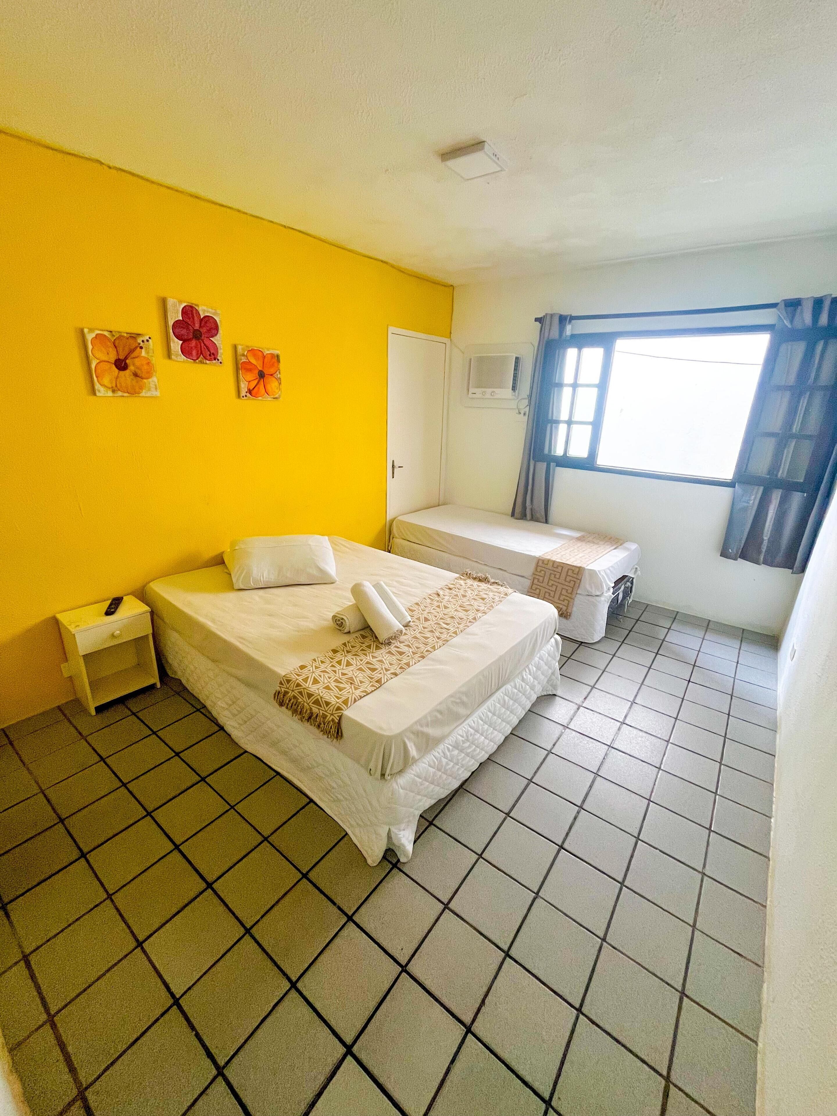 Quarto com uma cama grande, uma cama menor, uma mesa de cabeceira, uma janela com cortinas e um painel com três quadros de flores.