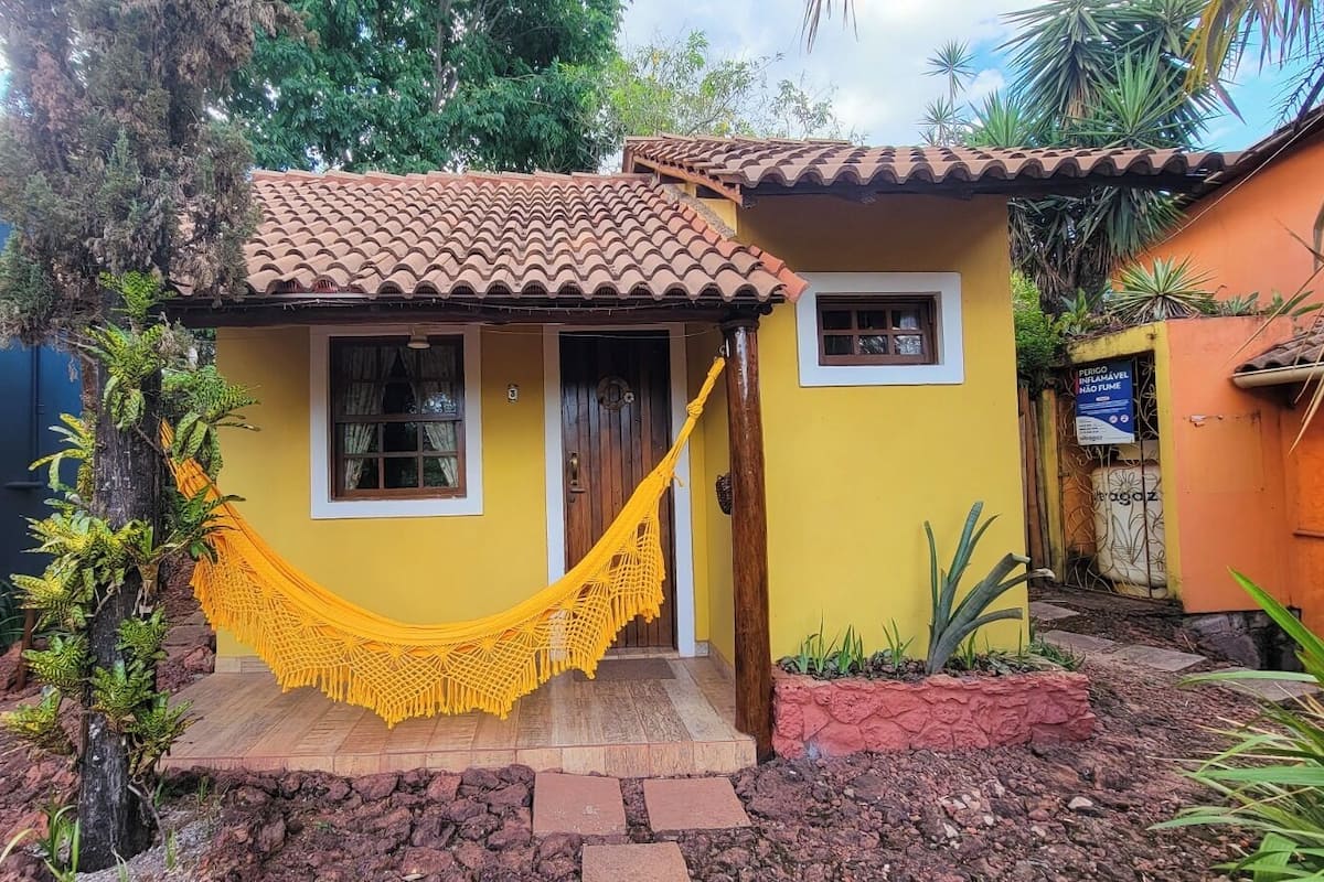 Uma casa amarela com telhado de telhas, uma rede e uma pequena varanda.