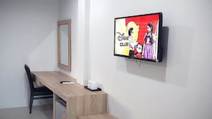 Quarto com escrivaninha de madeira, uma cadeira, televisão na parede exibindo 'Disney Club' e um pequeno armário.