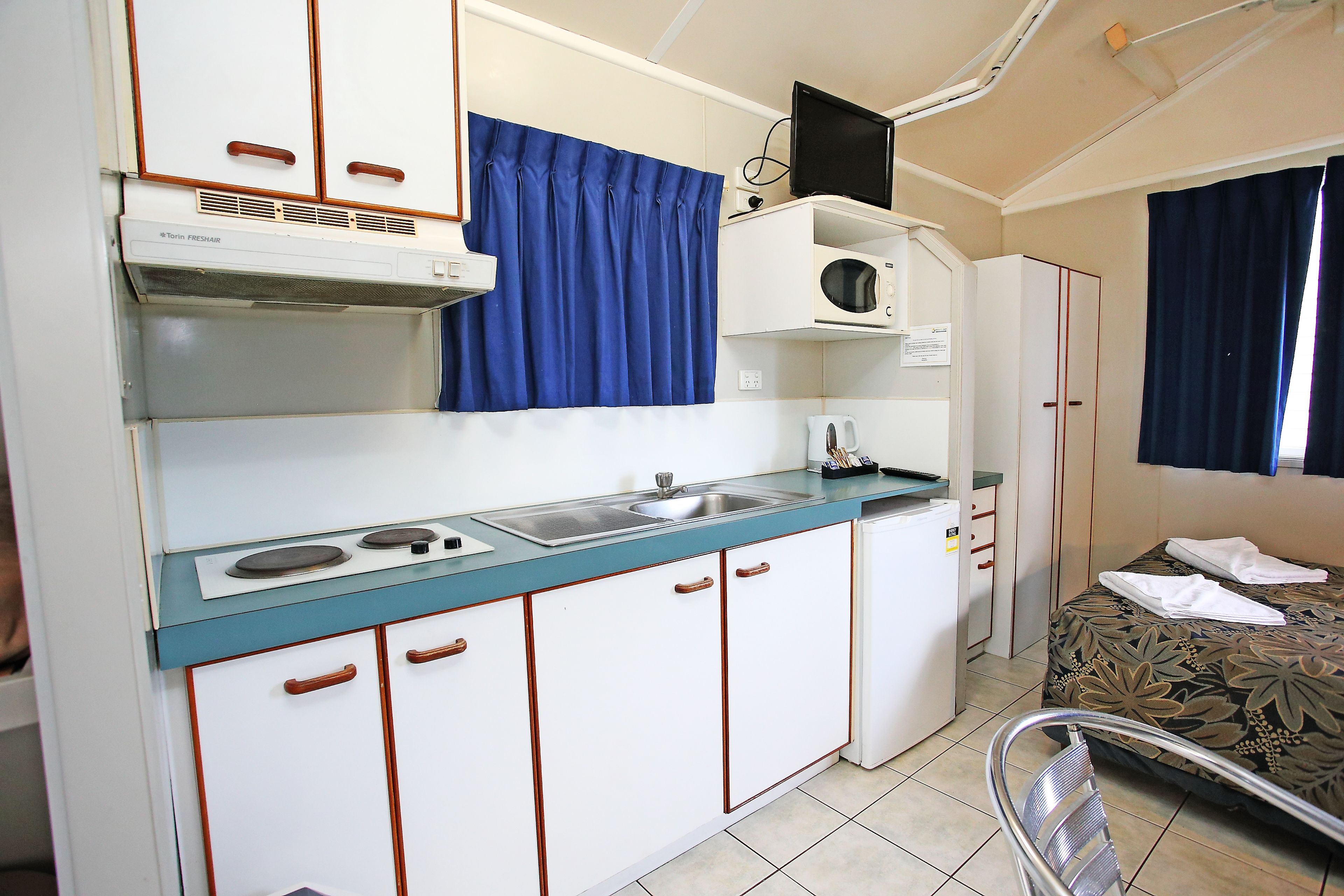 Cozinha compacta com micro-ondas, fogão e pia. Há uma cama com colcha estampada e uma janela com cortinas.