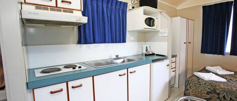 Cozinha compacta com micro-ondas, fogão e pia. Há uma cama com colcha estampada e uma janela com cortinas.