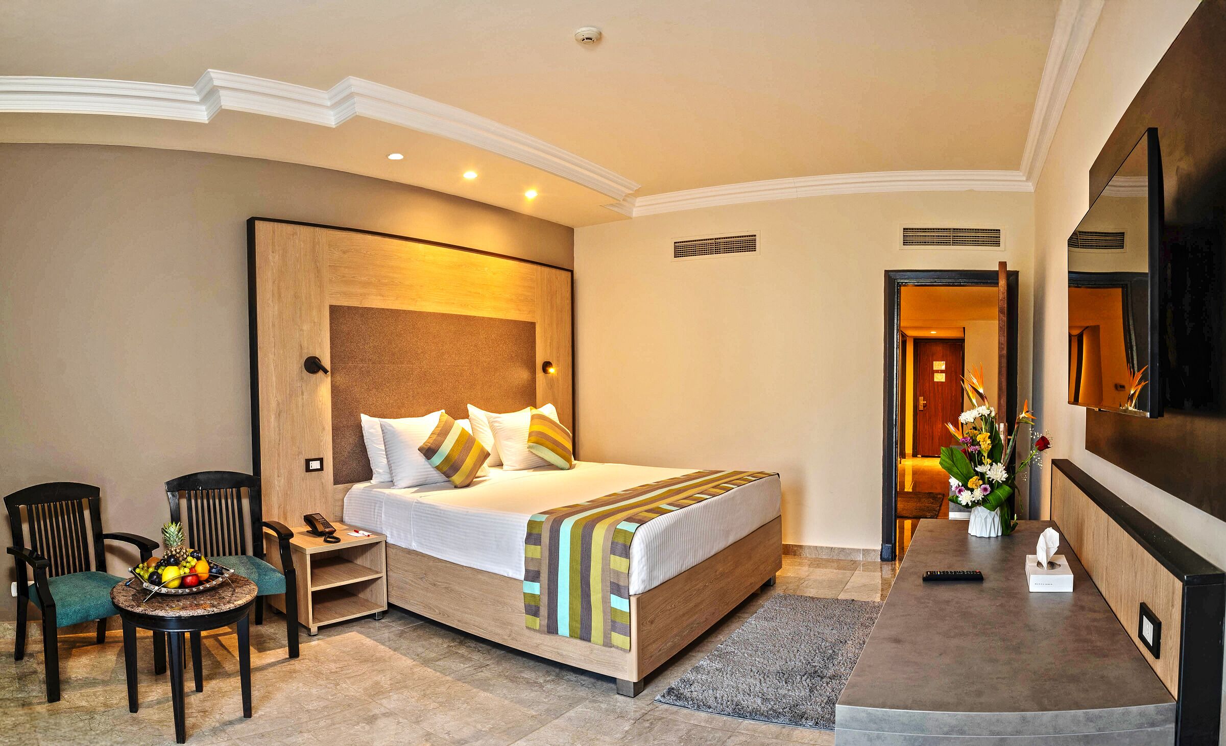 Quarto de hotel com uma cama grande, uma escrivaninha com telefone, uma cadeira, uma mesinha com frutas e um espelho na parede.