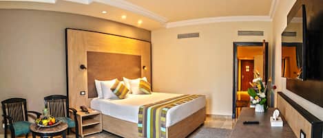Quarto de hotel com uma cama grande, uma escrivaninha com telefone, uma cadeira, uma mesinha com frutas e um espelho na parede.