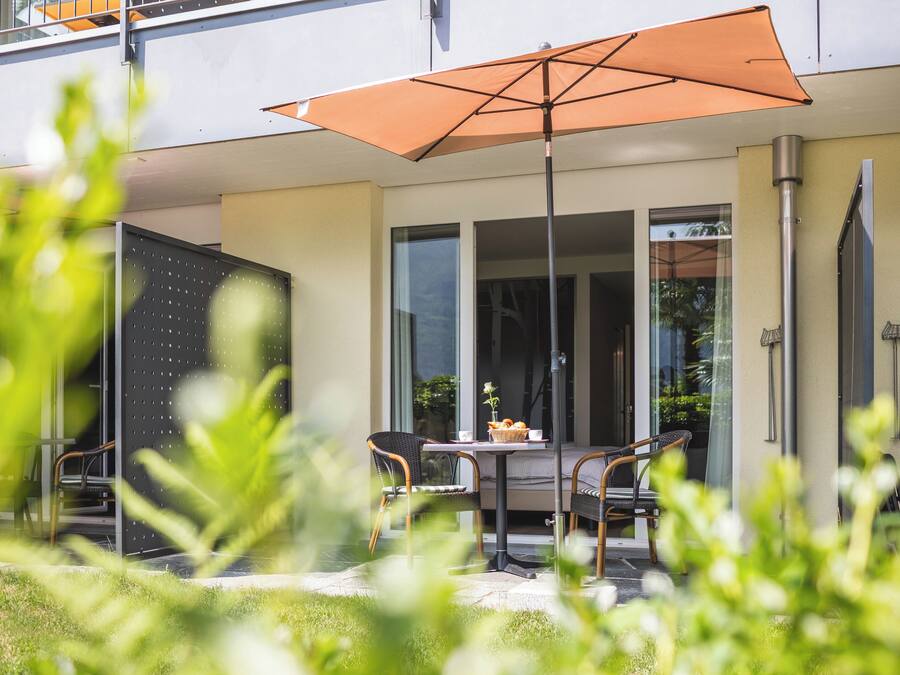 Um pátio externo com mesa e cadeiras, um guarda-sol e um edifício moderno ao fundo.