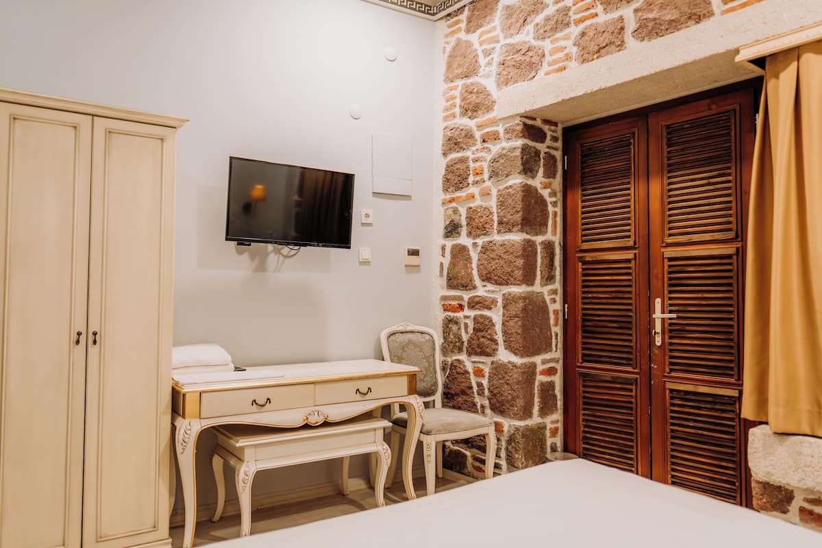 Quarto com parede de pedra, persianas de madeira, televisão, escrivaninha e uma cadeira.
