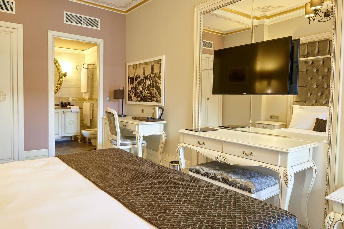 Quarto de hotel com cama, escrivaninha, cadeira, televisão e banheiro visível por uma porta aberta.