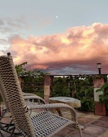 Uma cadeira de vime e uma mesa pequena em um alpendre ao ar livre, com vista para o céu e nuvens.
