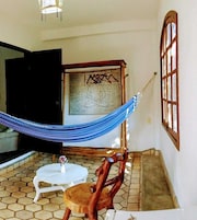 Quarto com rede, uma cadeira de madeira, uma mesa pequena e uma janela grande.