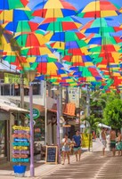 Rua repleta de guarda-chuvas coloridos, uma placa com diferentes opções de comida e pessoas caminhando.