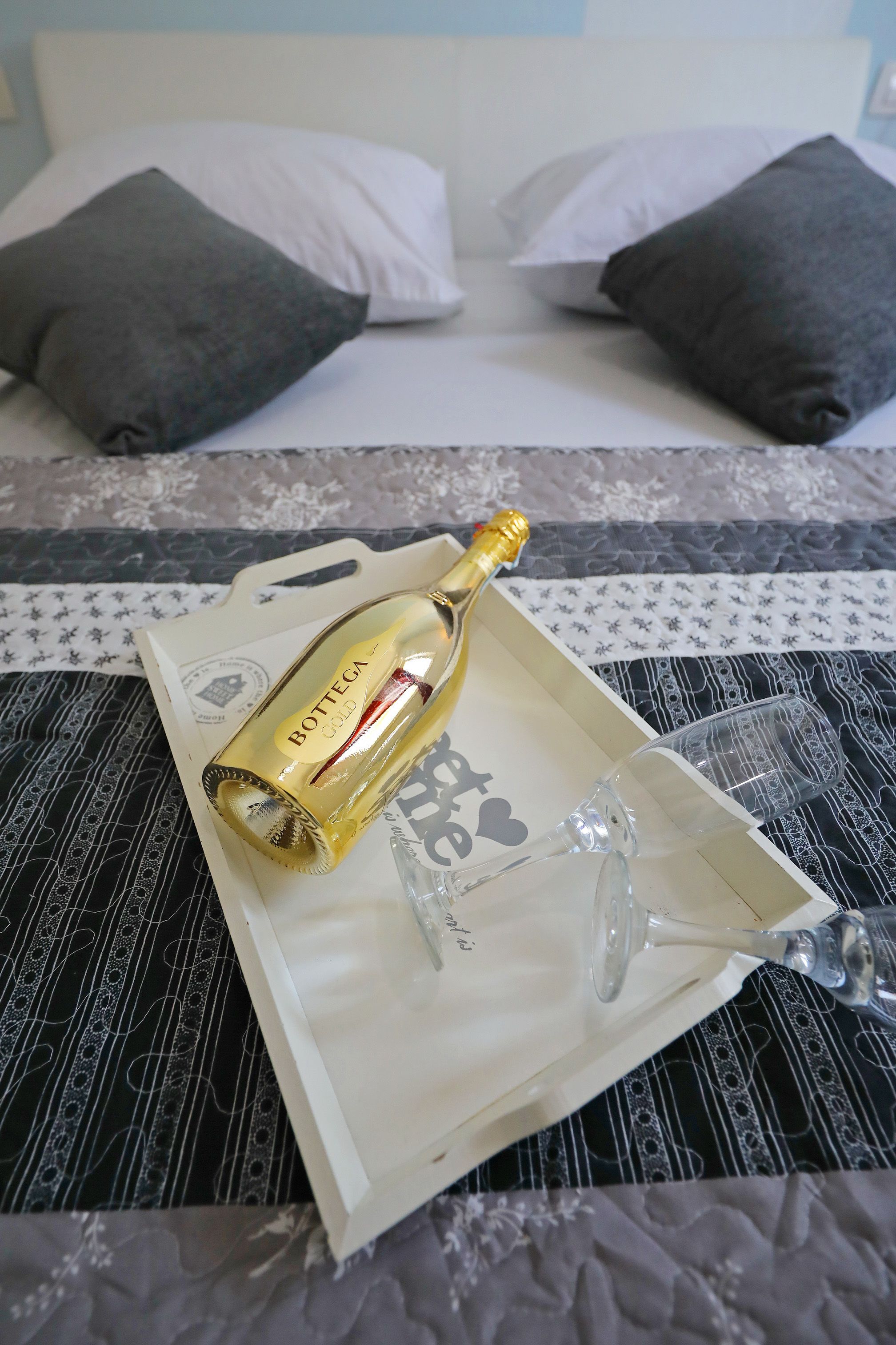 Uma garrafa de champanhe e uma taça em um tabuleiro sobre a cama, com roupa de cama e almofadas estampadas.