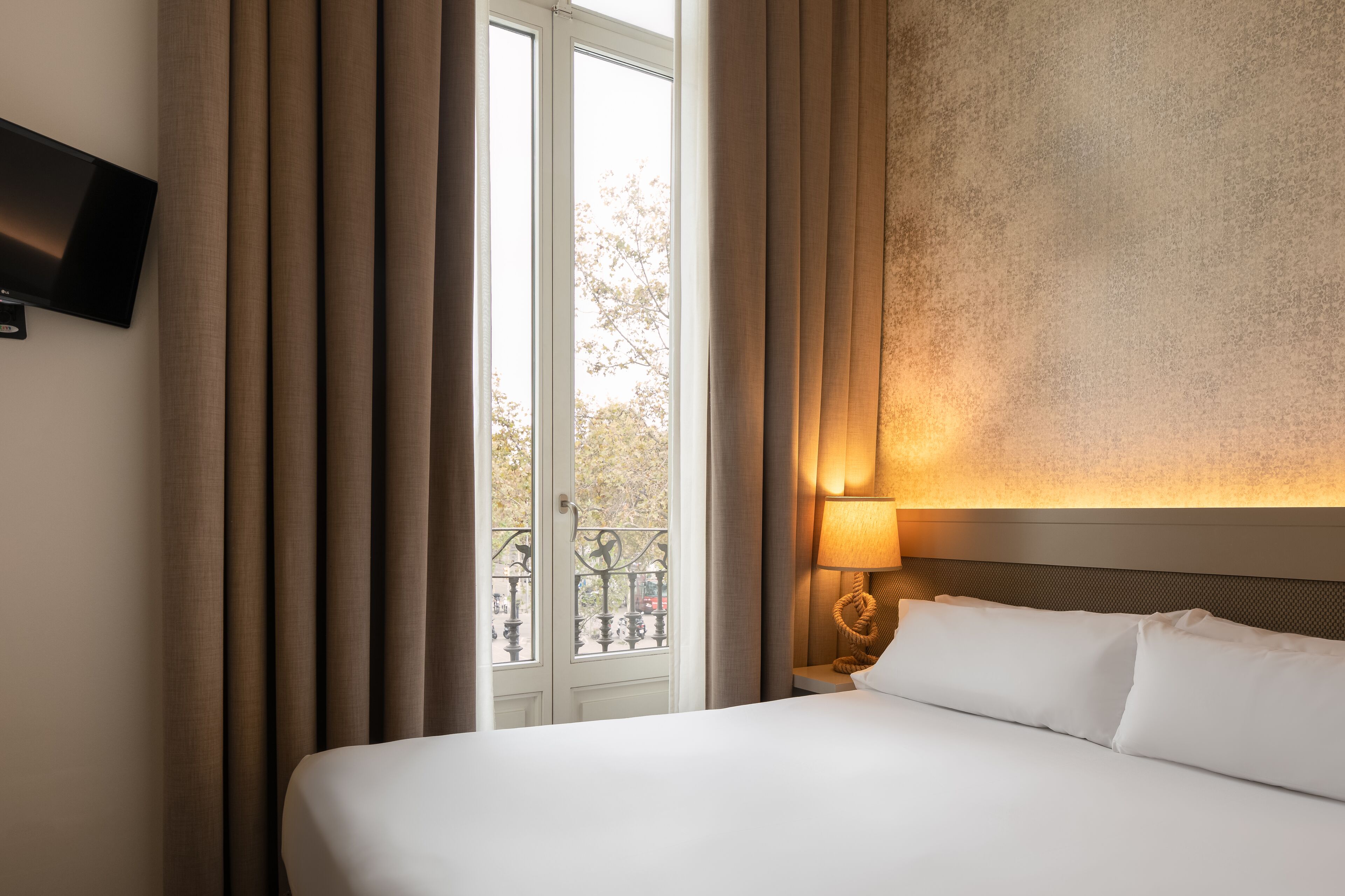 Quarto de hotel com cama, cortinas, televisão presa à parede, janela com vista para árvores e varanda, além de luminária na parede.
