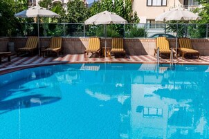 Piscina com espreguiçadeiras e guarda-sóis, rodeada por um deck ladrilhado e vegetação.