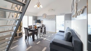 Uma sala de estar moderna com um sofá cinzento, uma área de jantar com mesa e cadeiras, uma escada com guarda-corrimão de metal e um candelabro.
