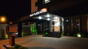 Exterior do Hotel Biazi Plaza, com entrada bem iluminada, uma placa de identificação e área ajardinada com plantas.