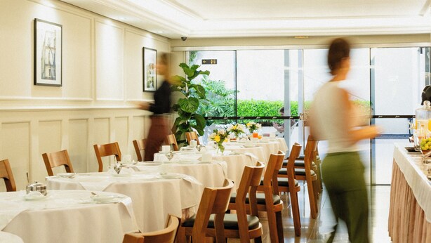 Restaurante com mesas postas para refeição, cadeiras de madeira e uma pessoa caminhando.