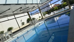 Piscina interior moderna com paredes de vidro transparente, cadeiras brancas e plantas em vasos.