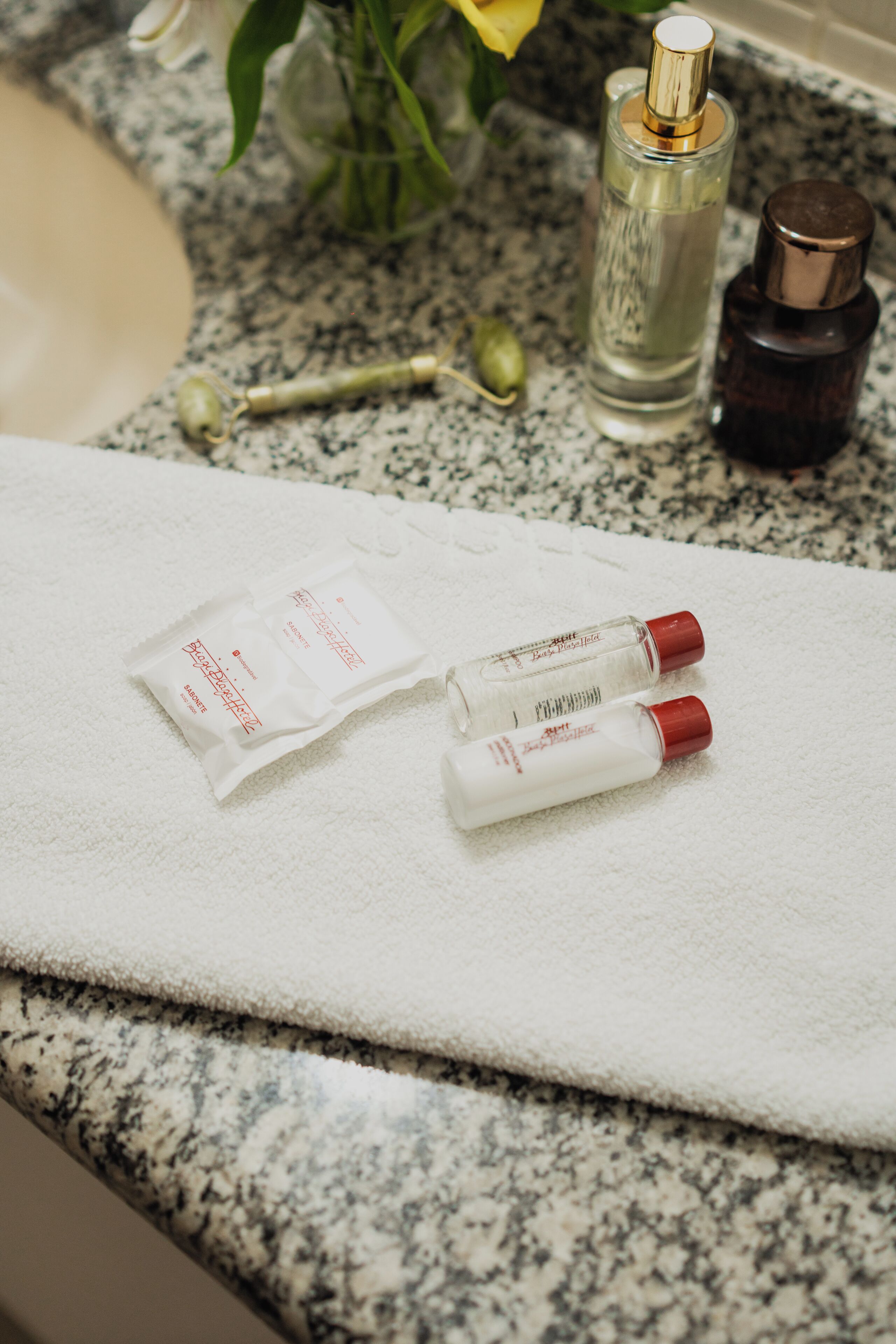 Uma bancada de banheiro com uma toalha, duas embalagens de loção, um frasco de perfume e um pequeno vaso com uma flor amarela.