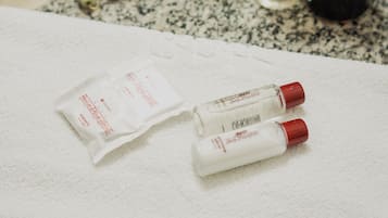 Uma bancada de banheiro com uma toalha, duas embalagens de loção, um frasco de perfume e um pequeno vaso com uma flor amarela.