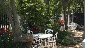 Um pátio com jardim, mobiliado com cadeiras e mesas de metal branco, rodeado por vegetação exuberante e plantas floridas.