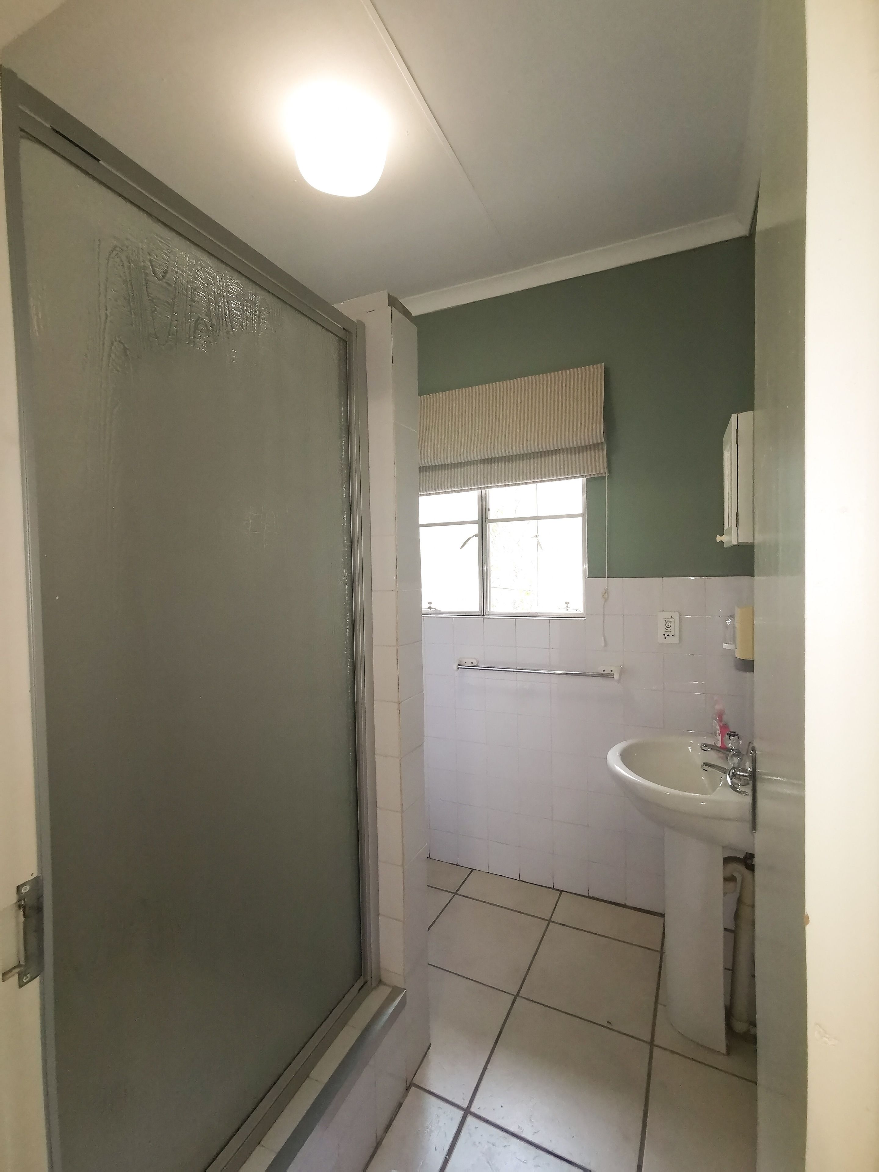 Um banheiro com chuveiro, pia, janela com persianas e porta.
