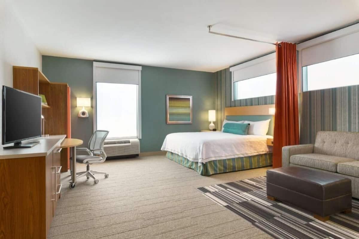 Quarto de hotel com cama, escrivaninha com televisão, poltrona, sofá e uma grande janela.
