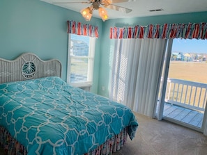 Quarto com cama, ventoinha no teto, janela com persianas e vista para a praia.