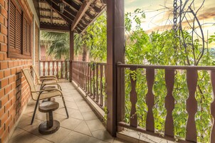 Varanda coberta com cadeiras de vime, uma mesa pequena e vista para o verde e um edifício ao fundo.