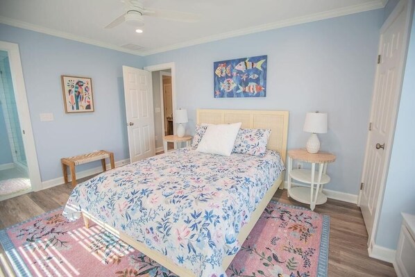 Quarto com colcha florida, mesa de cabeceira com abajur, um banquinho e um quadro na parede.
