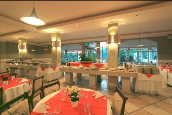 Salão de festas com mesas redondas, toalhas brancas e detalhes em vermelho.