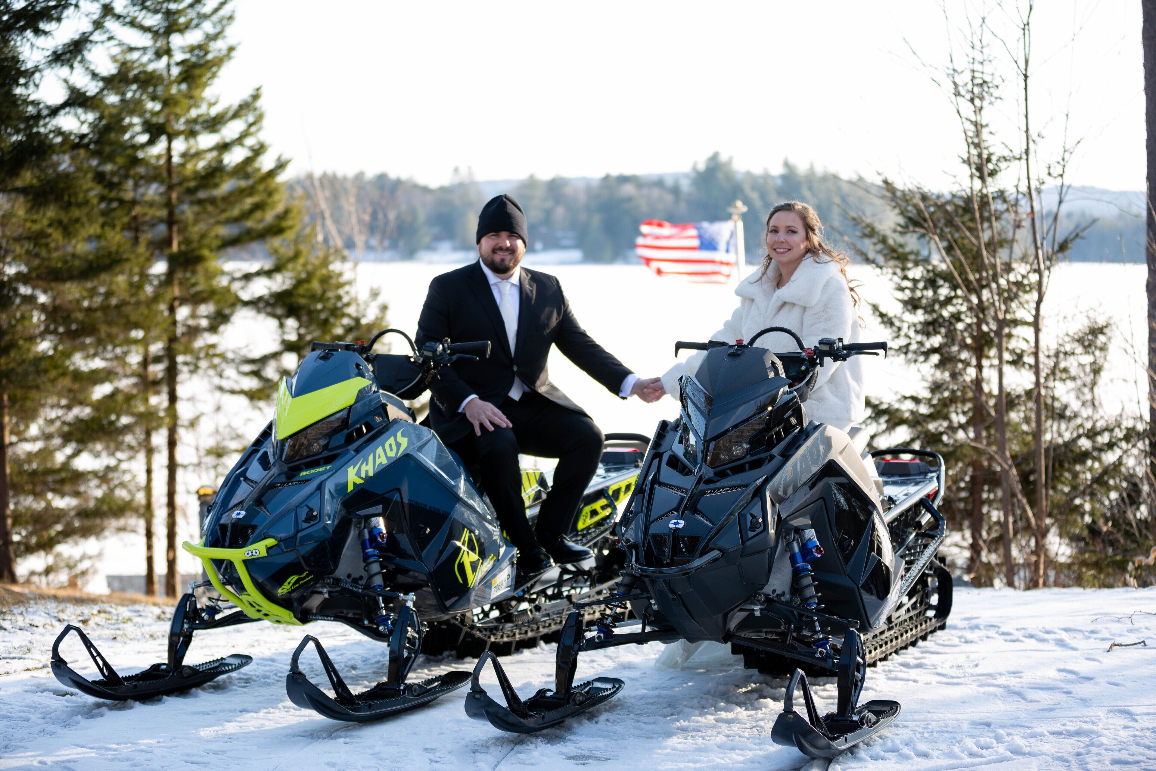 Duas pessoas em motos de neve, com um lago e árvores ao fundo.