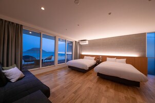 Quarto de hotel com duas camas, um sofá, uma janela grande com vista para o mar e piso de madeira.