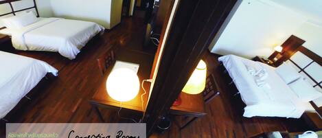 Quarto de hotel com duas camas de solteiro, piso de madeira, uma mesa com abajur e uma pequena escrivaninha com cadeira.