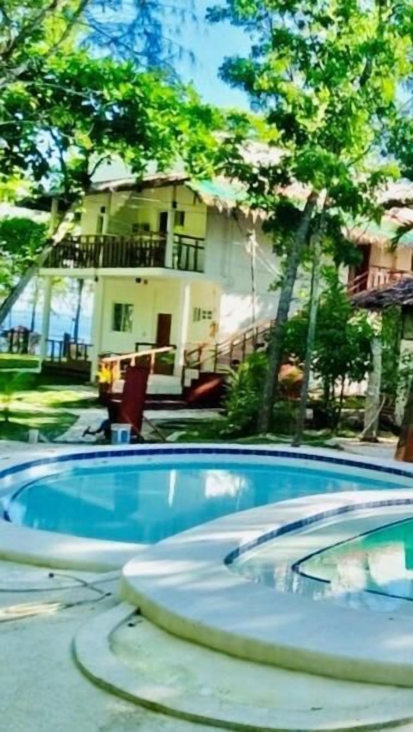 Uma piscina com água azul cristalina, cercada por vegetação exuberante e um edifício com varanda.